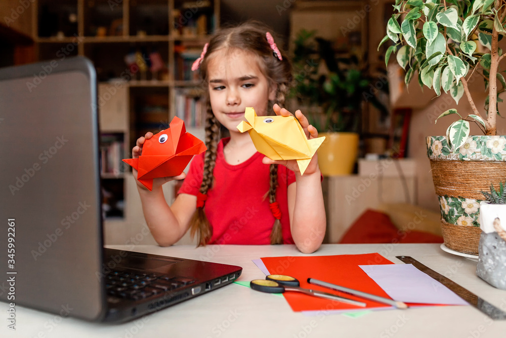 Girl doing origami fish with color paper looking video on laptop ...