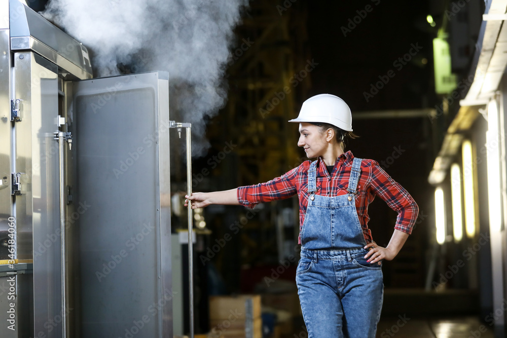 Young girl in a work dress and white hard hat in a factory. Woman in a ...