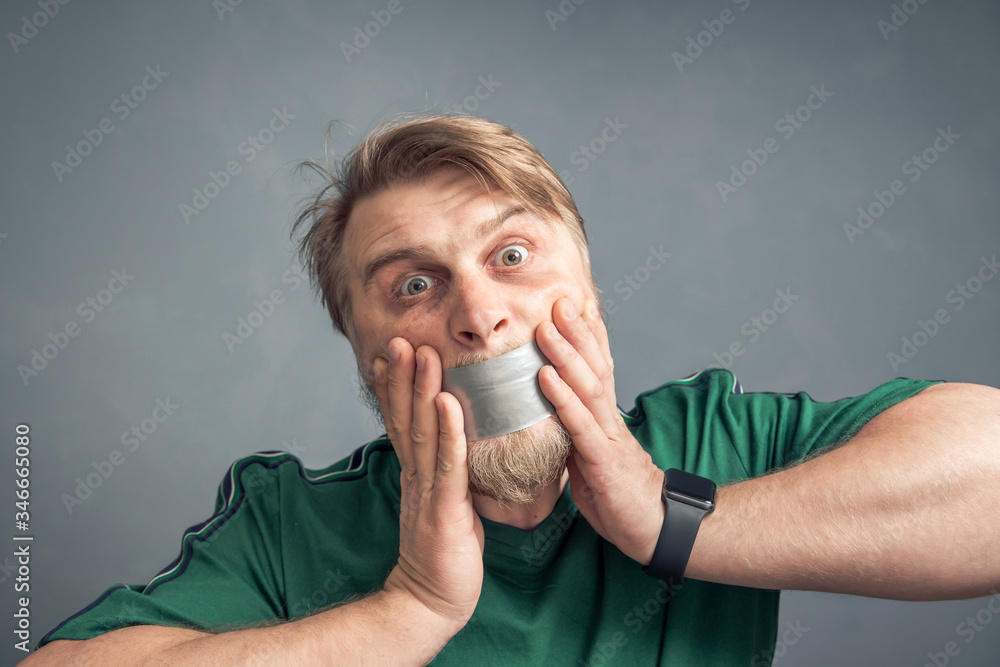 A close-up portrait of a bearded man with his mouth taped shut, holding ...