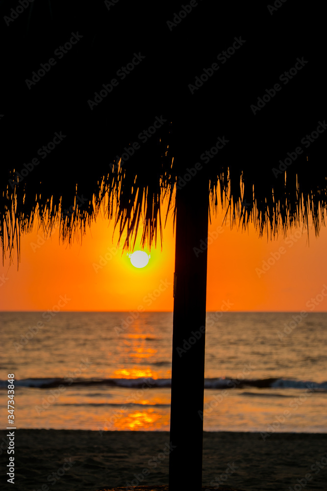Sol ocultándose en la playa detrás de una palapa Stock Photo | Adobe Stock