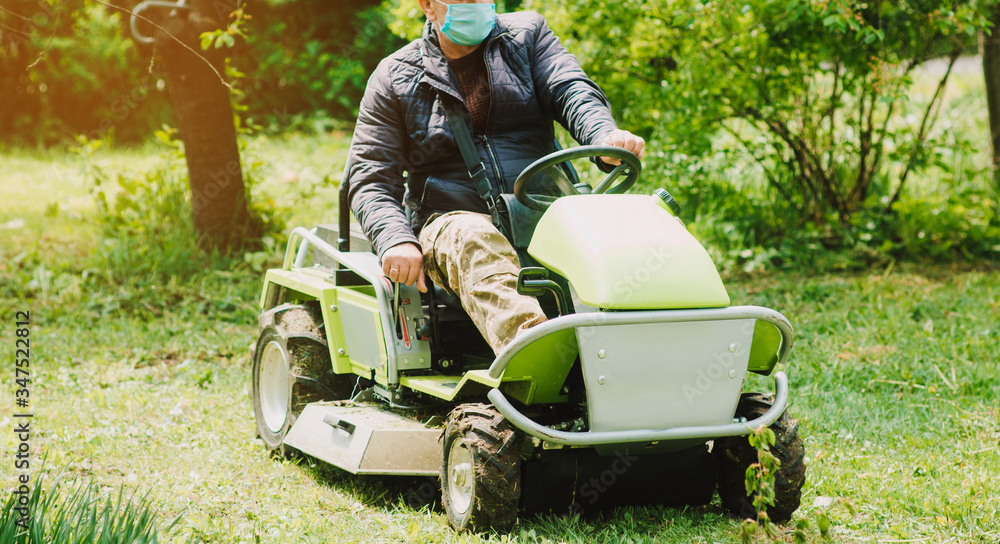 Senior gardener in medical mask driving a riding lawn mower in a garden ...