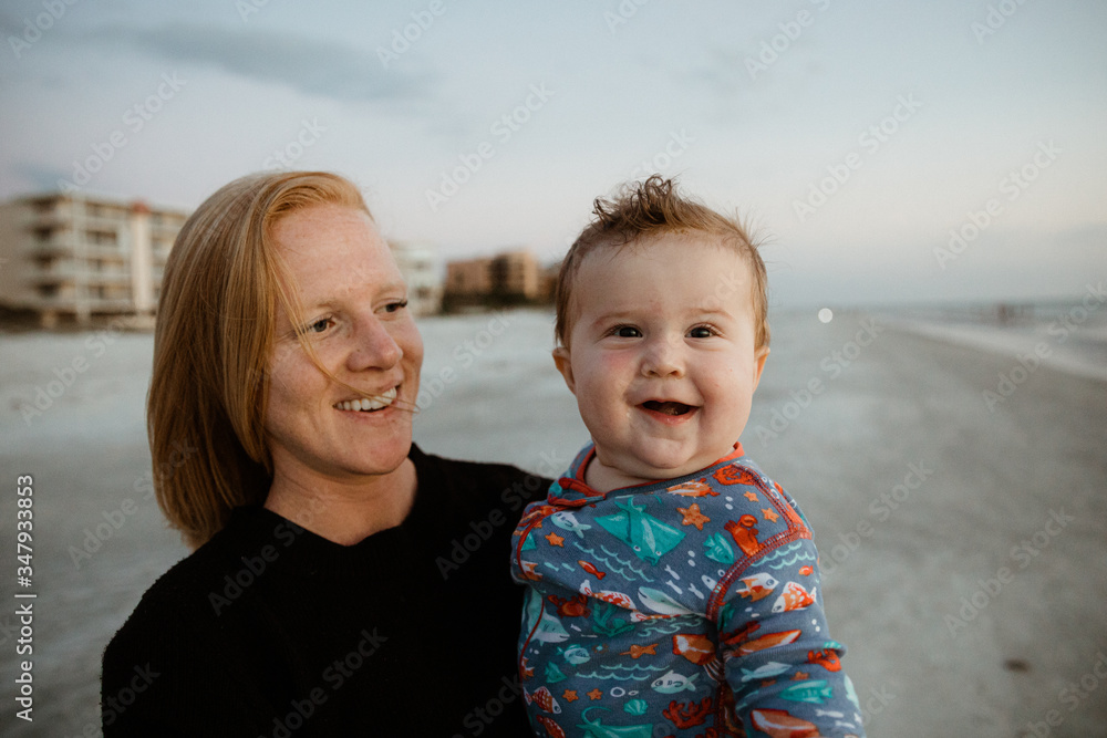 fat baby boy with crooked smile held by young red haired mom on beach ...