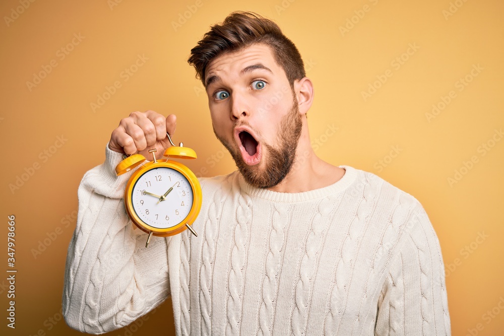 Young blond man with beard and blue eyes holding alarm clock over ...