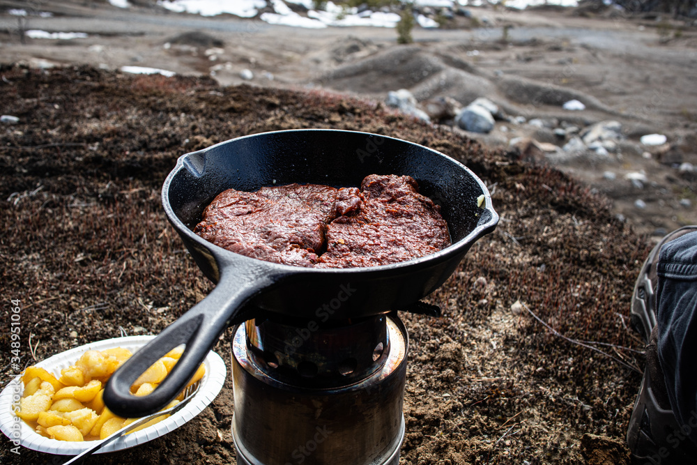 Cooking steaks and potatoes with cast iron frying pan next to sand pit ...