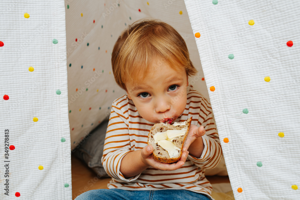 Cute little boy eating bread and butter in a hut, looking up at a ...