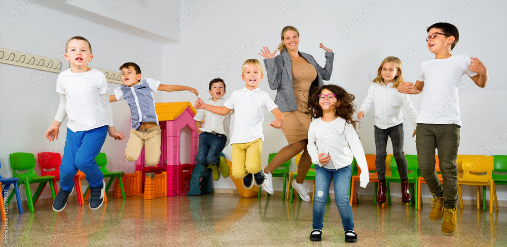 Positive female teacher and group of schoolkids jumping in classroom ...