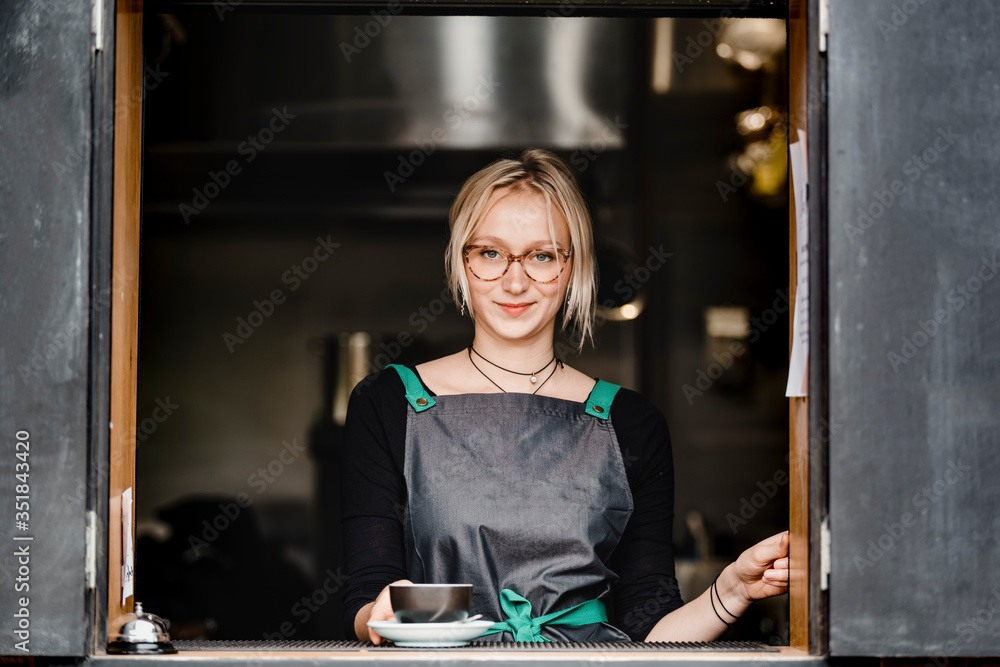 A beautiful blonde young barista girl serving a cup of coffee in tiny ...
