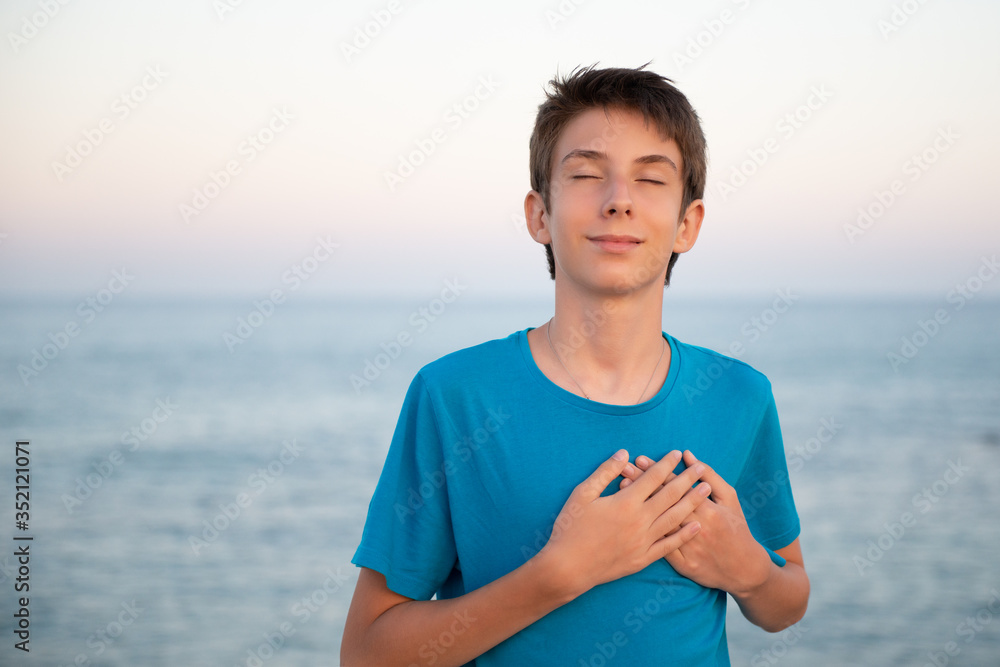 Handsome young happy boy with eyes closed at beach holds hands on chest ...
