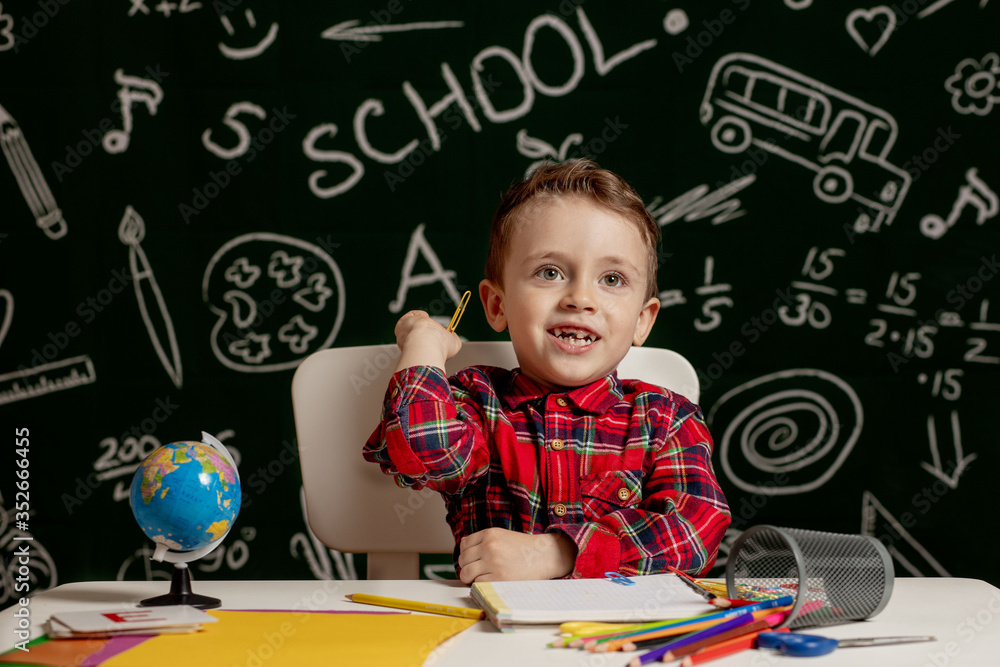 Emotional school boy sitting on the desk with many school supplies ...