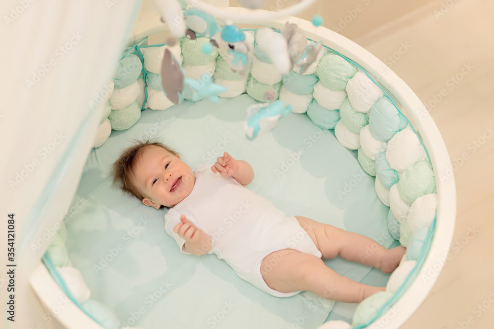 Happy baby boy lies on his back in a white round bed in the bedroom ...