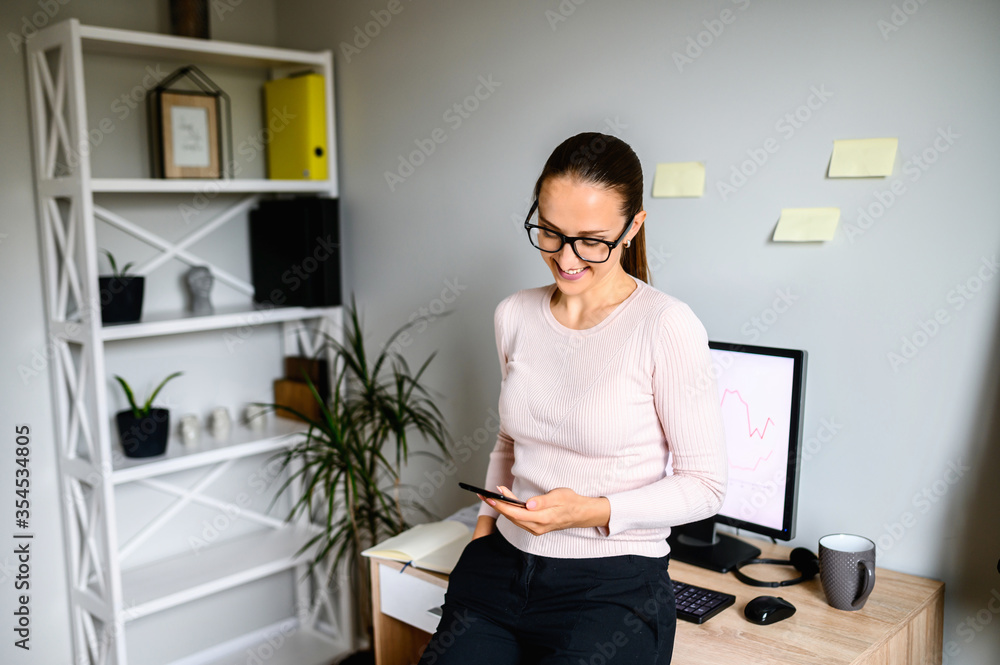Smiling woman looks at the phone screen and standing in home office ...