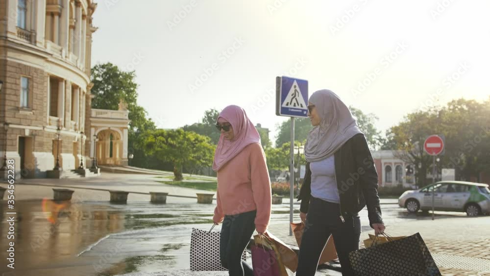 Two muslim women in casual clothes, sunglasses and hijabs. Smiling and talking, walking with ...