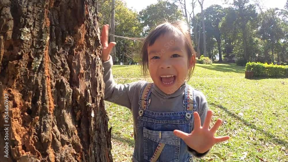 Cheerful child girl playing peekaboo behind a tree in the park during ...