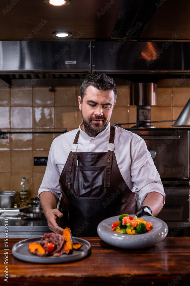 Handsome chef in leather apron standing at counter and serving dishes ...