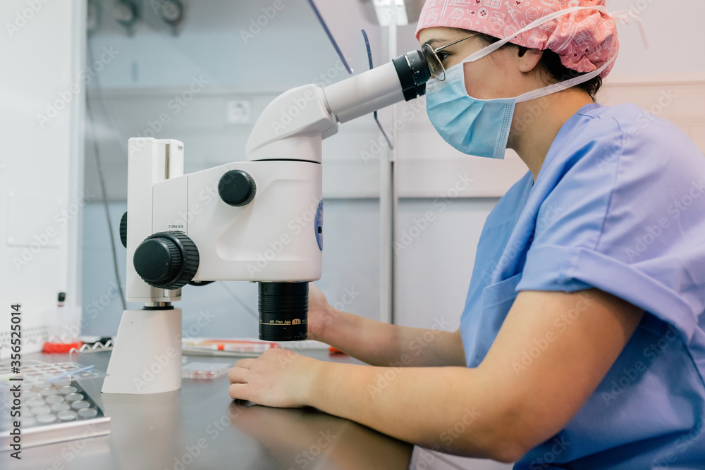 Side view of woman in medical uniform and mask using modern microscope ...