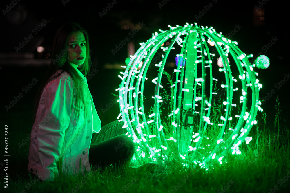 The girl at night sits near a glowing ball of green color. A glowing ...