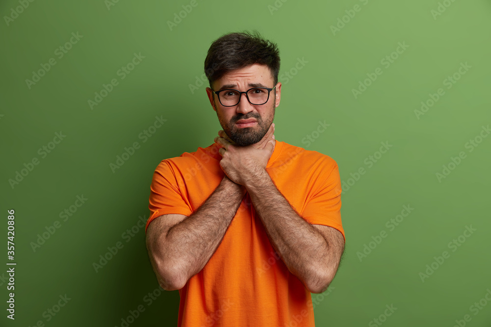 Frustrated unshaven man keeps hands on neck, suffers from sore throat ...