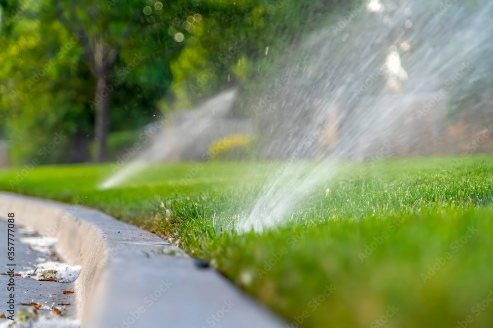 Curb of the paved road with water sprinklers on the vivid green grassy ...