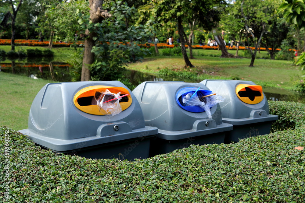 Row of plastic bins behind bush trees in park. Three bins are used for ...