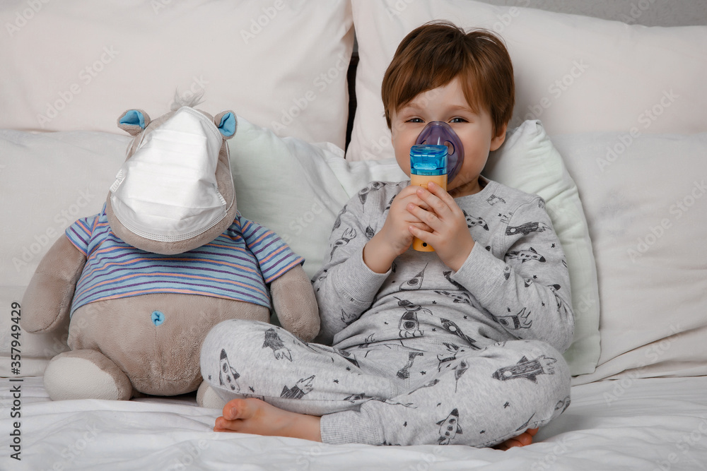 beautiful boy is sitting with a toy on the bed in an inhalation mask ...