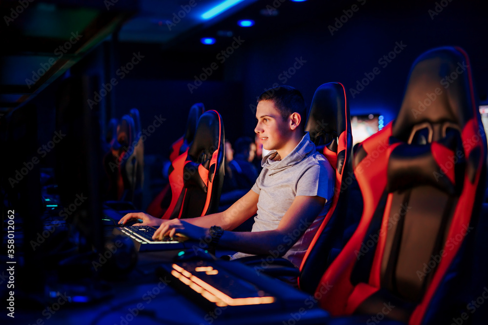 Smiling young professional gamer playing video game at internet cafe or ...