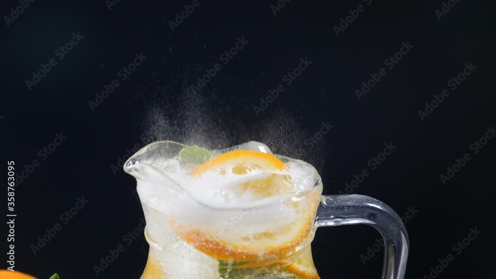 Close-up shot of aerated cold water being poured in transparet jar with sliced orange and ice ...