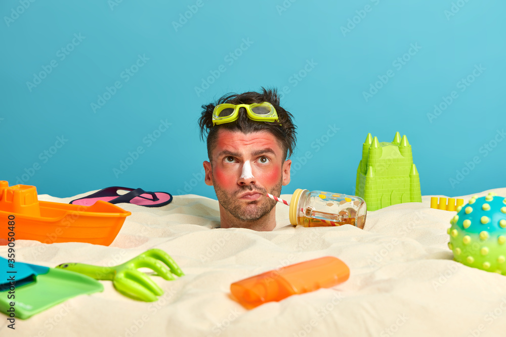 Thoughtful young man buried in sand, drinks cold refreshing beverage ...