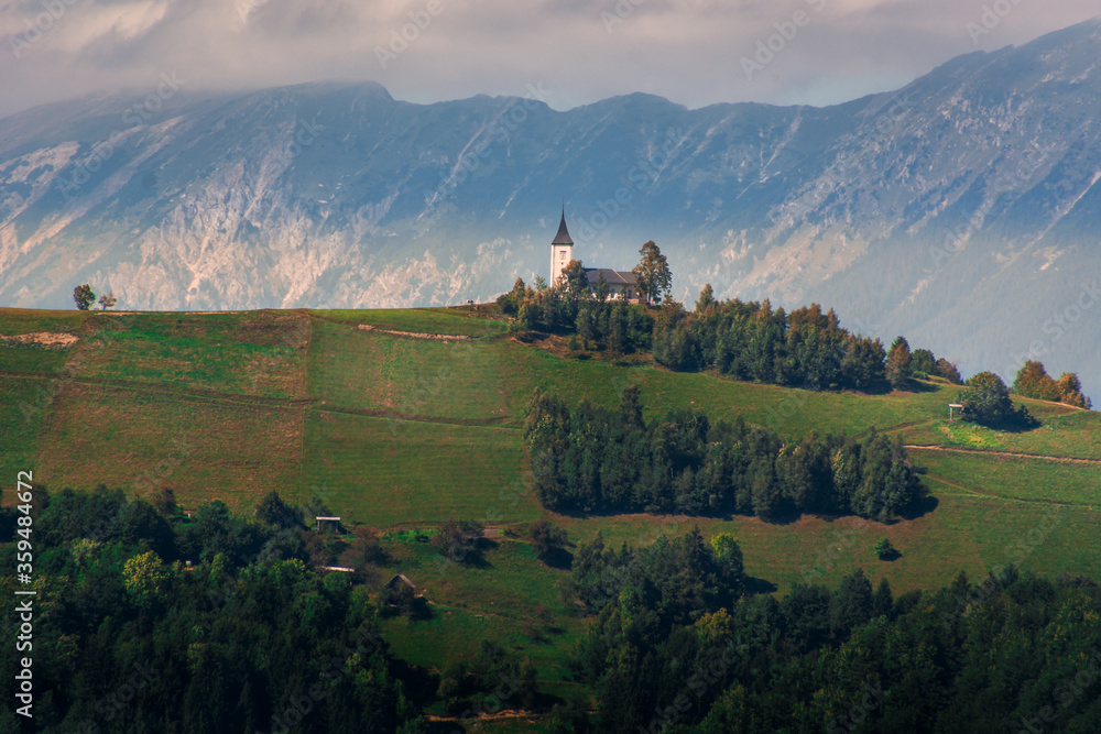 Jamnik church on hilltop with Alps mountains in background. Hill slope ...