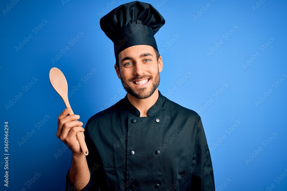 Young cooker man with beard wearing uniform holding wooden spoon over ...