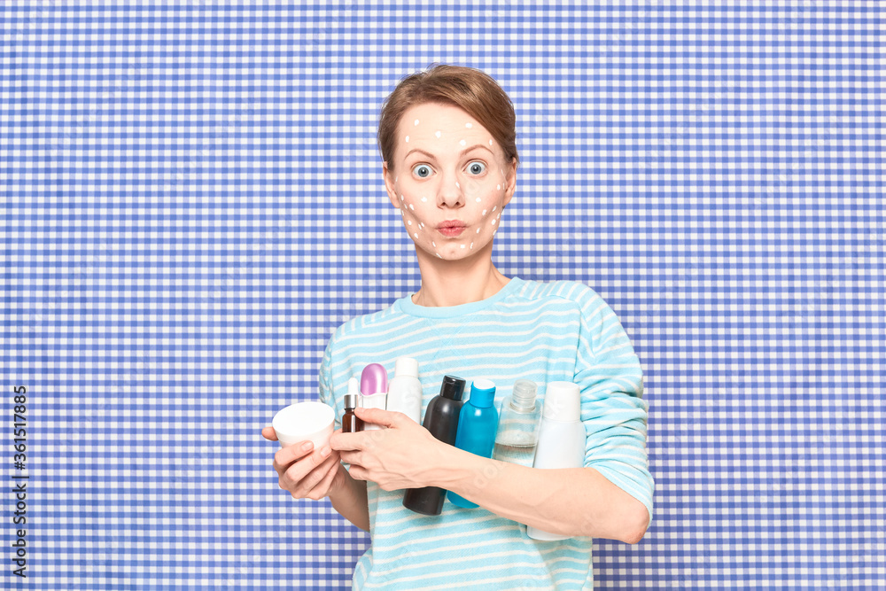 Surprised girl with problem face skin is holding many cosmetic bottles ...