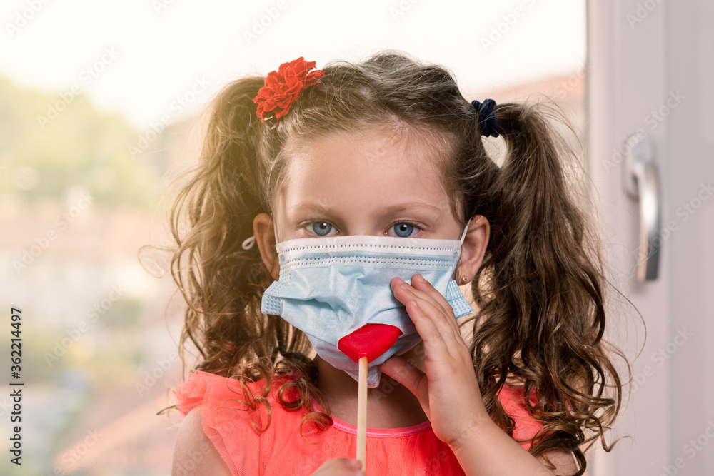 cute little girl wearing a surgical mask is eating a lollipop through a ...