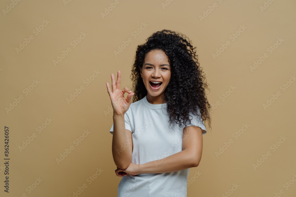 Half length shot of joyful curly young woman makes okay gesture, enjoys ...