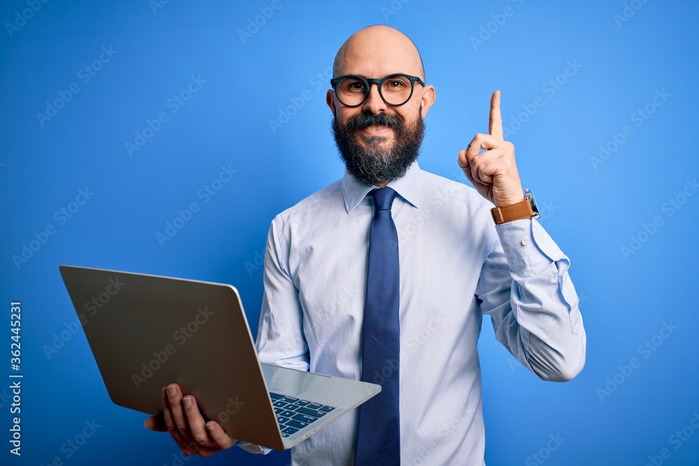 Handsome bald business man with beard working using laptop over blue ...
