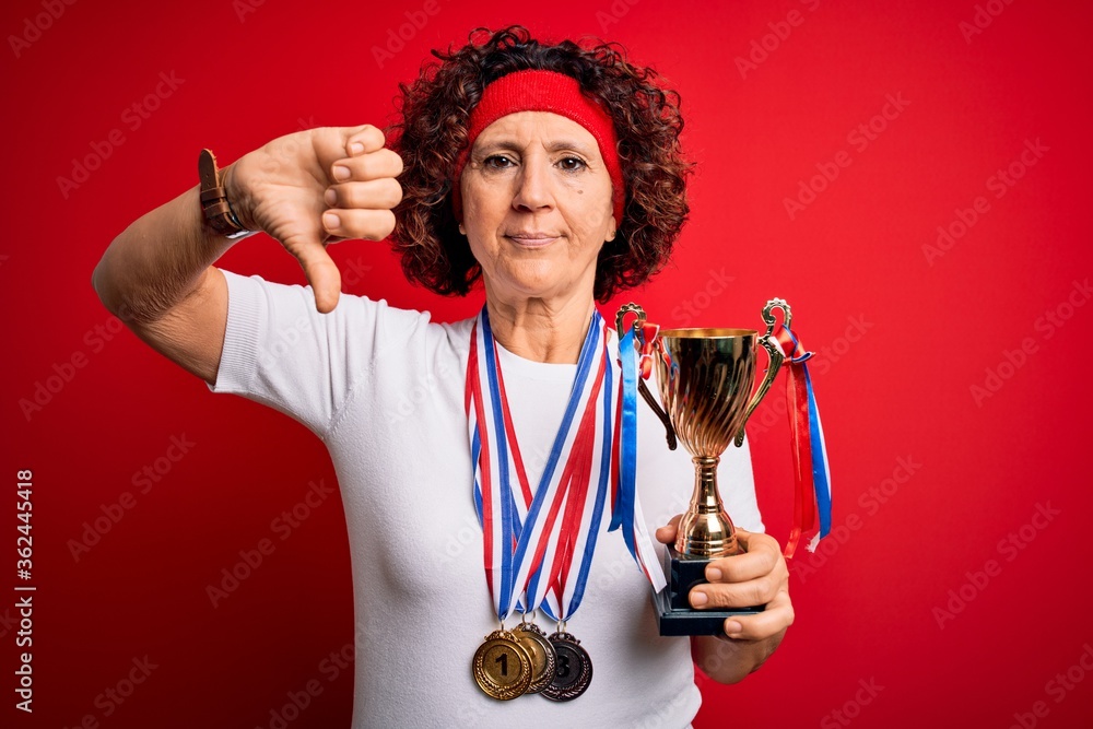 Middle age curly woman winning medals holding trophy over isolated red ...