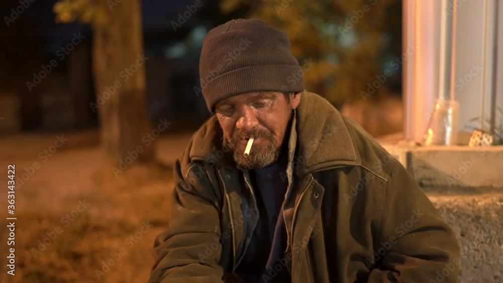 Homeless sick man smoking cigarette on the street at night. Lonely cold ...