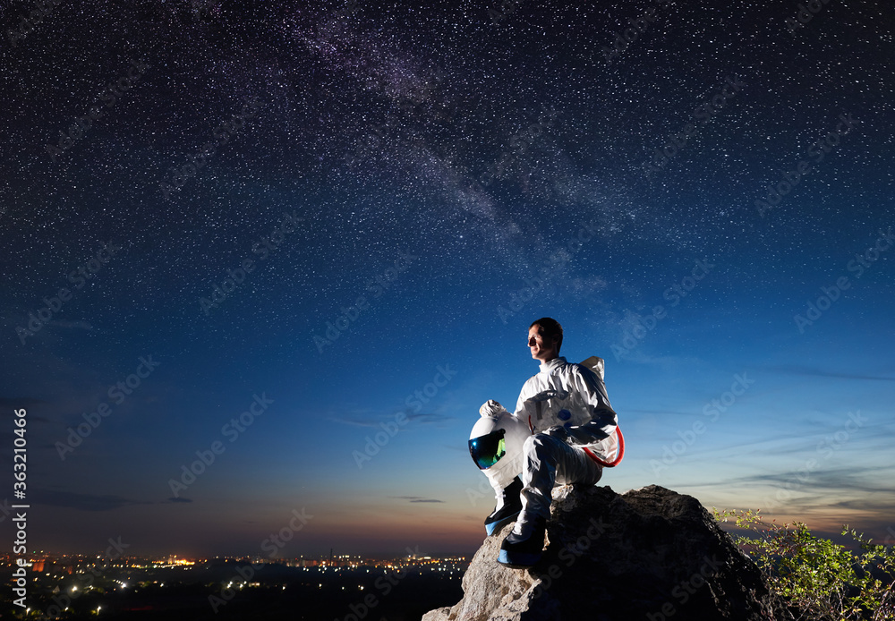Side view of astronaut sitting on top of rocky hill and looking at ...
