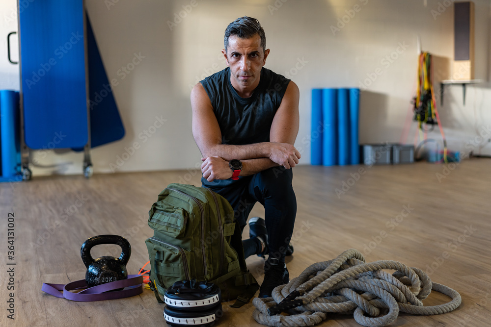 Motivated boot camp instructor kneeling with gym equipment in gym hall ...