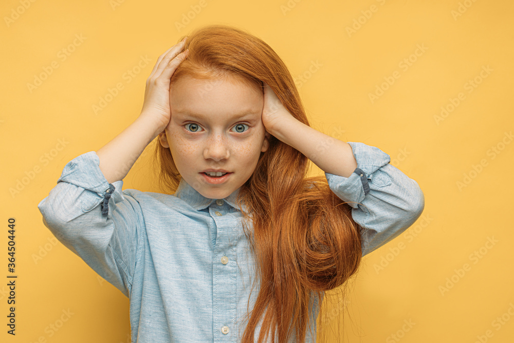 little girl at a loss, perplexed girl with red hair touch her head and look at camera. in shock ...