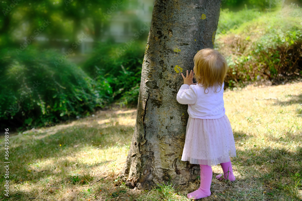 little kid, a girl with blonde hair, stands near a tree in the park ...