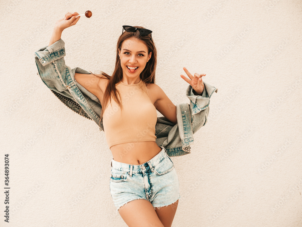 Portrait of beautiful cute model in summer hipster jeans jacket clothes. Girl eating chupa chups ...