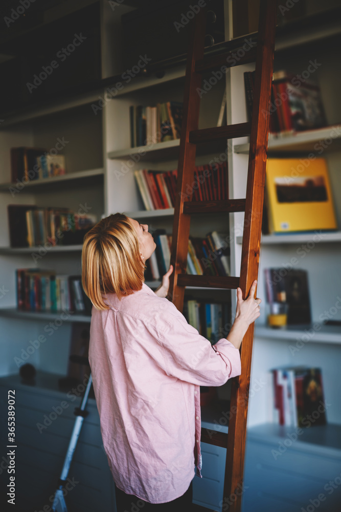 Young blonde woman climbing rolling library ladder to take some book ...