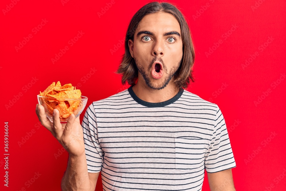 Young handsome man holding nachos potato chips scared and amazed with ...