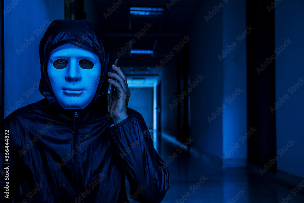 Anonymous computer hacker in white mask and hoodie. Obscured dark face calling on cellphone on ...