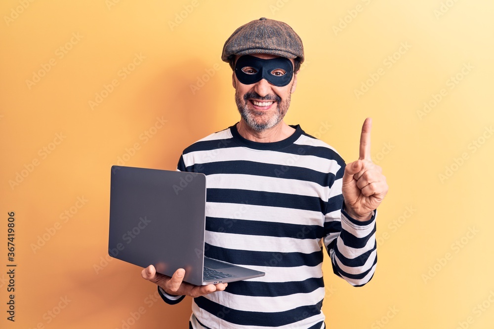 Middle age handsome burglar man wearing cap and mask using laptop over ...