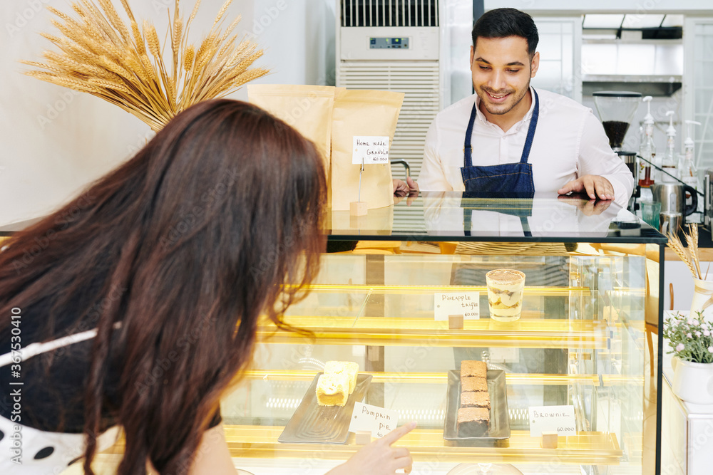 Young woman choosing dessert in showcase of bakery and asking waiter to ...