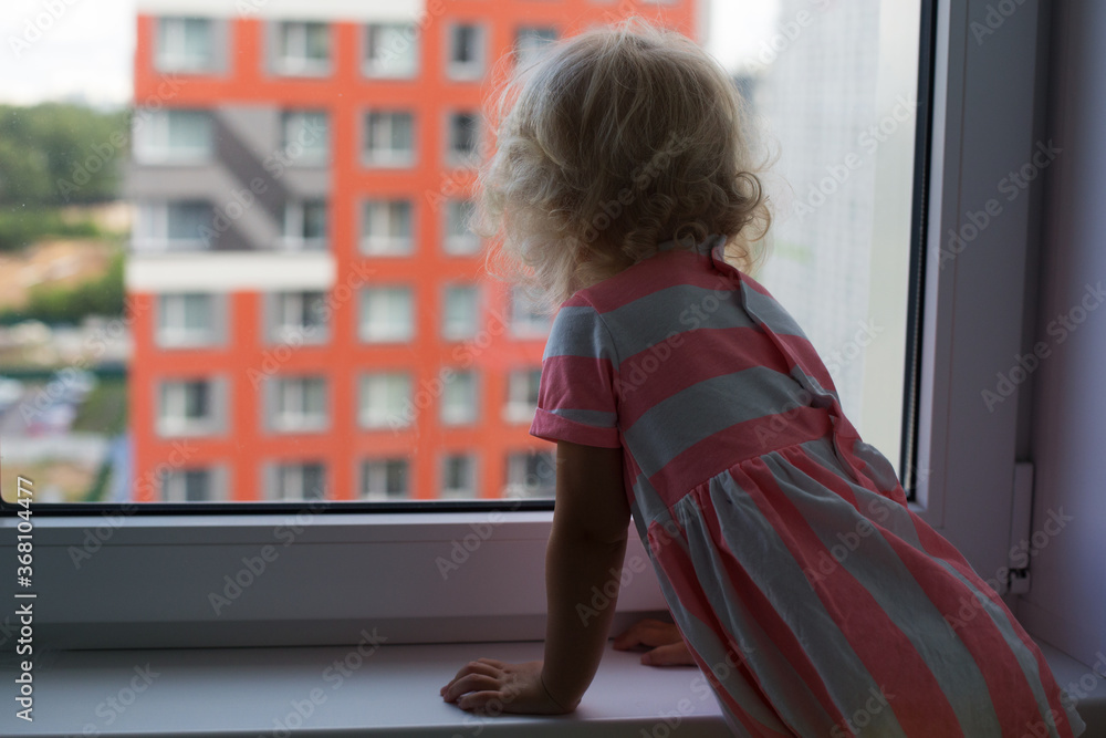 Small curly girl with blond hair looking outside from the window in ...