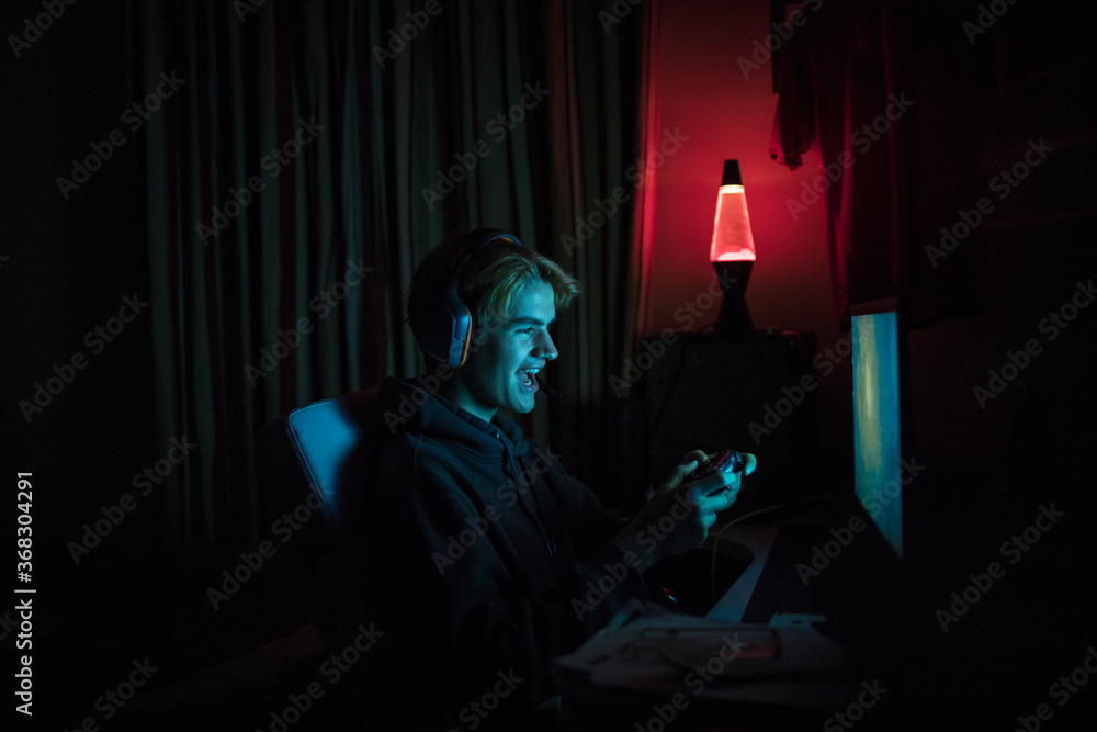 Smiling teenage boy playing video game at computer in dark bedroom ...