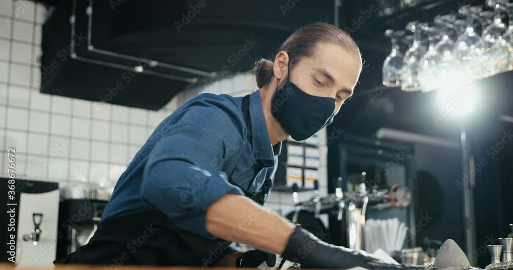Caucasian male barman in mask and gloves wiping surface of bar counter ...