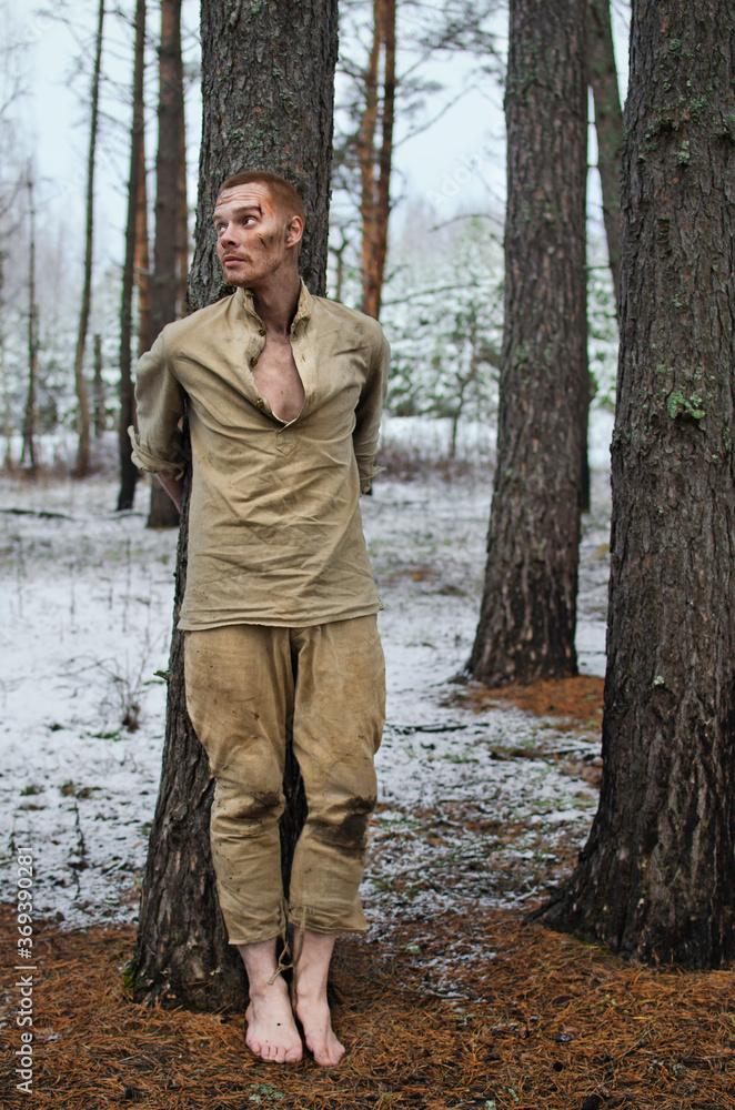 young man in military uniform tied to the trunk of an old tree barefoot ...