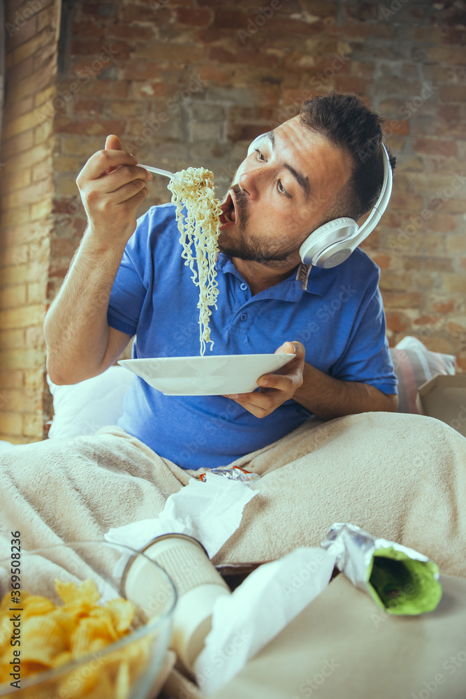 Passioned eating instant noodle. Lazy man living in his bed surrounded ...